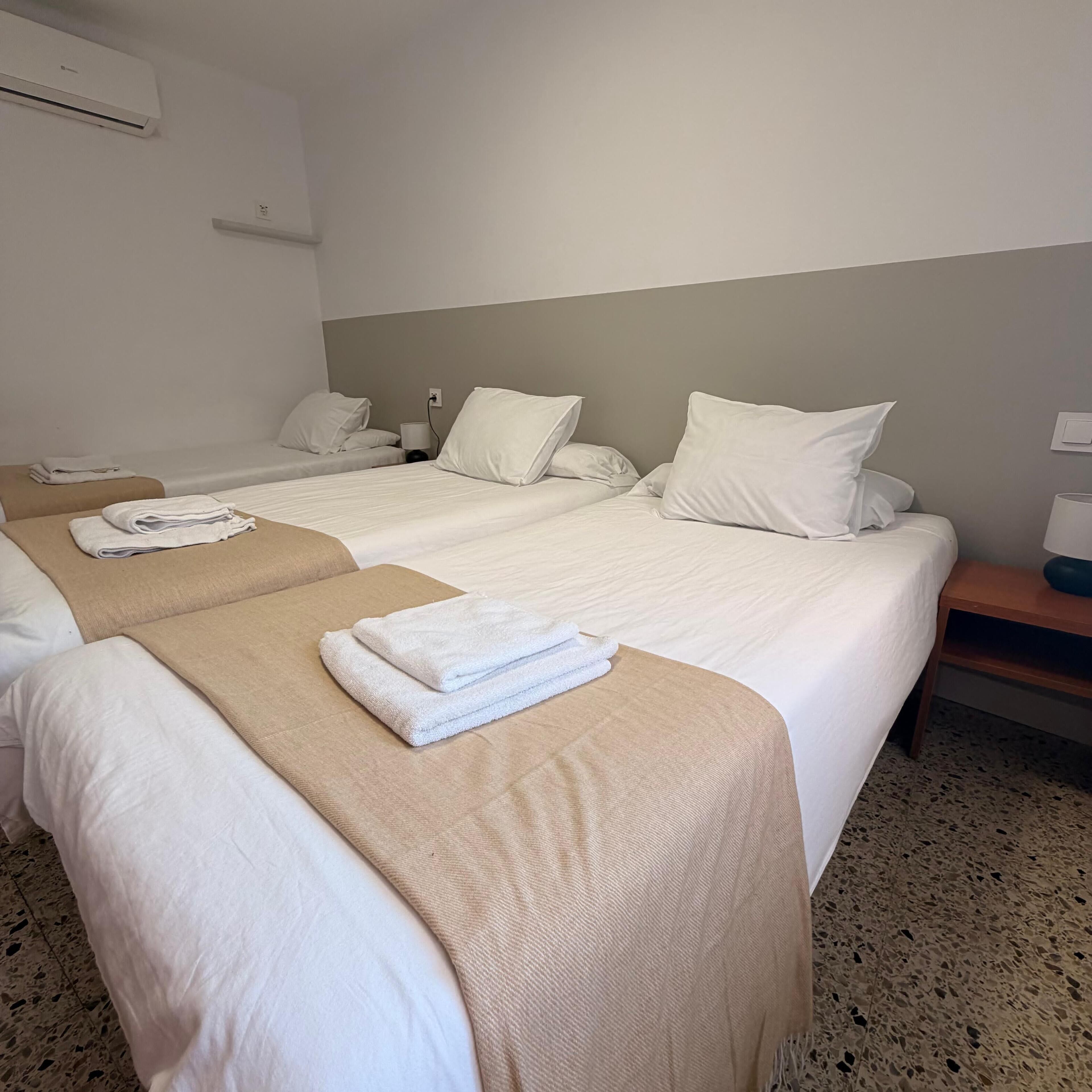 Quarto de hotel com duas camas, cada uma com lençóis brancos e colchas bege. Há uma pequena mesa com uma luminária e uma tomada na parede.
