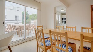 Uma área de refeições com uma mesa grande e cadeiras, uma porta de vidro deslizante que dá para uma varanda externa, e uma cozinha visível através de uma porta aberta.