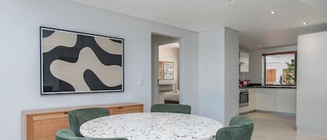 Uma zona de refeições moderna com uma mesa redonda e quatro cadeiras verdes, um aparador de vime e um grande quadro abstrato na parede.