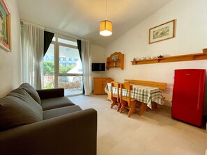 Uma sala de estar com sofá, mesa de jantar com cadeiras, geladeira vermelha e uma janela com cortinas.