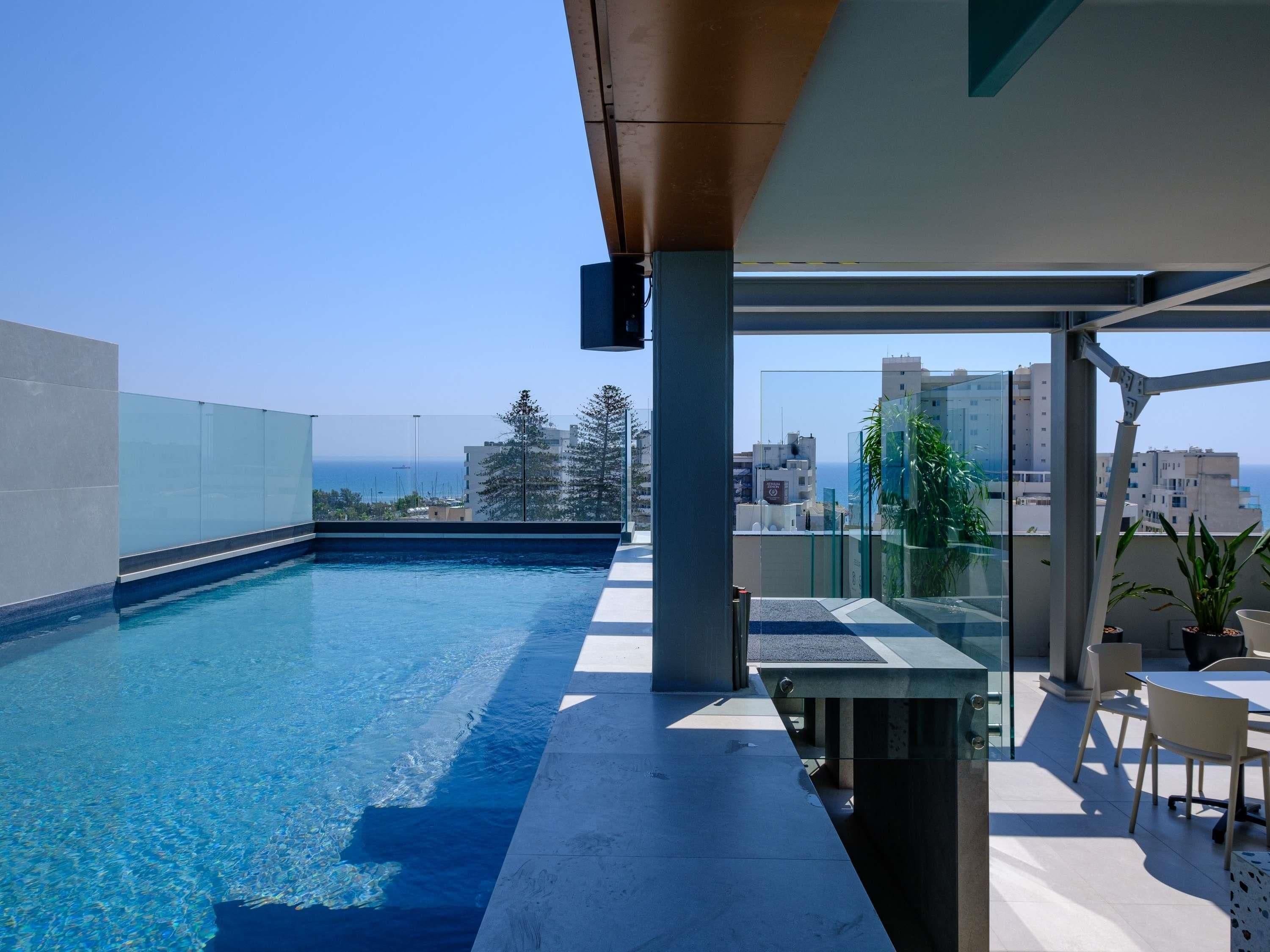 Piscina no terraço com água azul cristalina, guarda-corpo de vidro e vista para o mar e edifícios.