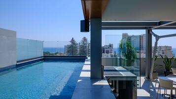 Piscina no terraço com água azul cristalina, guarda-corpo de vidro e vista para o mar e edifícios.