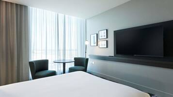 Quarto de hotel com uma cama grande, duas cadeiras, uma mesa pequena, uma televisão fixada na parede e uma janela com cortinas.