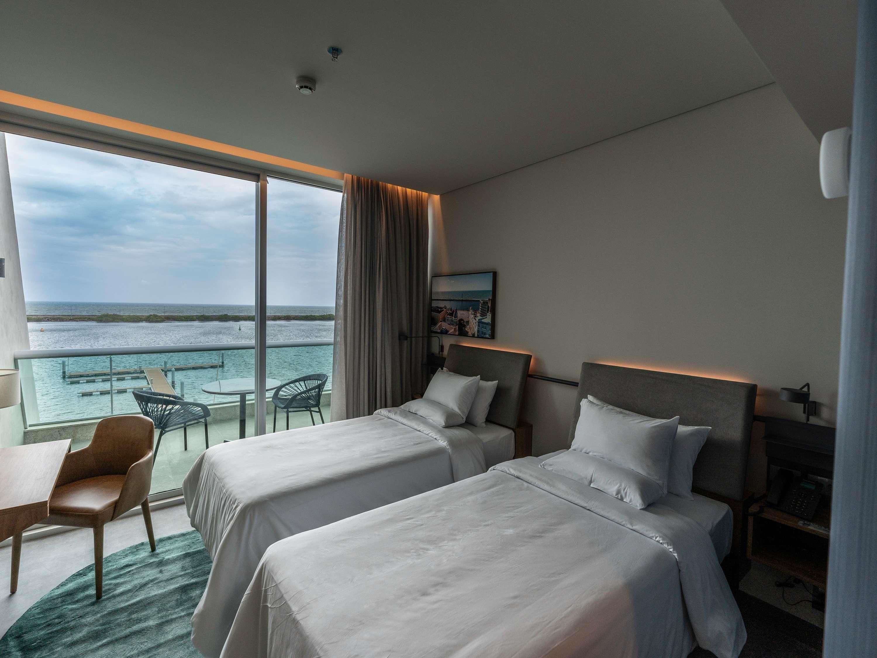 Quarto de hotel com duas camas, uma varanda com vista para o mar, área de refeições e uma pequena mesa.