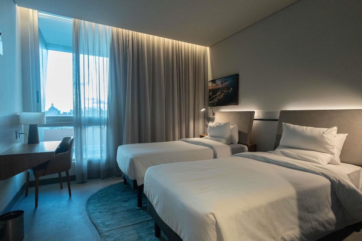 Quarto de hotel com duas camas, uma escrivaninha, uma cadeira e uma janela grande com cortinas.