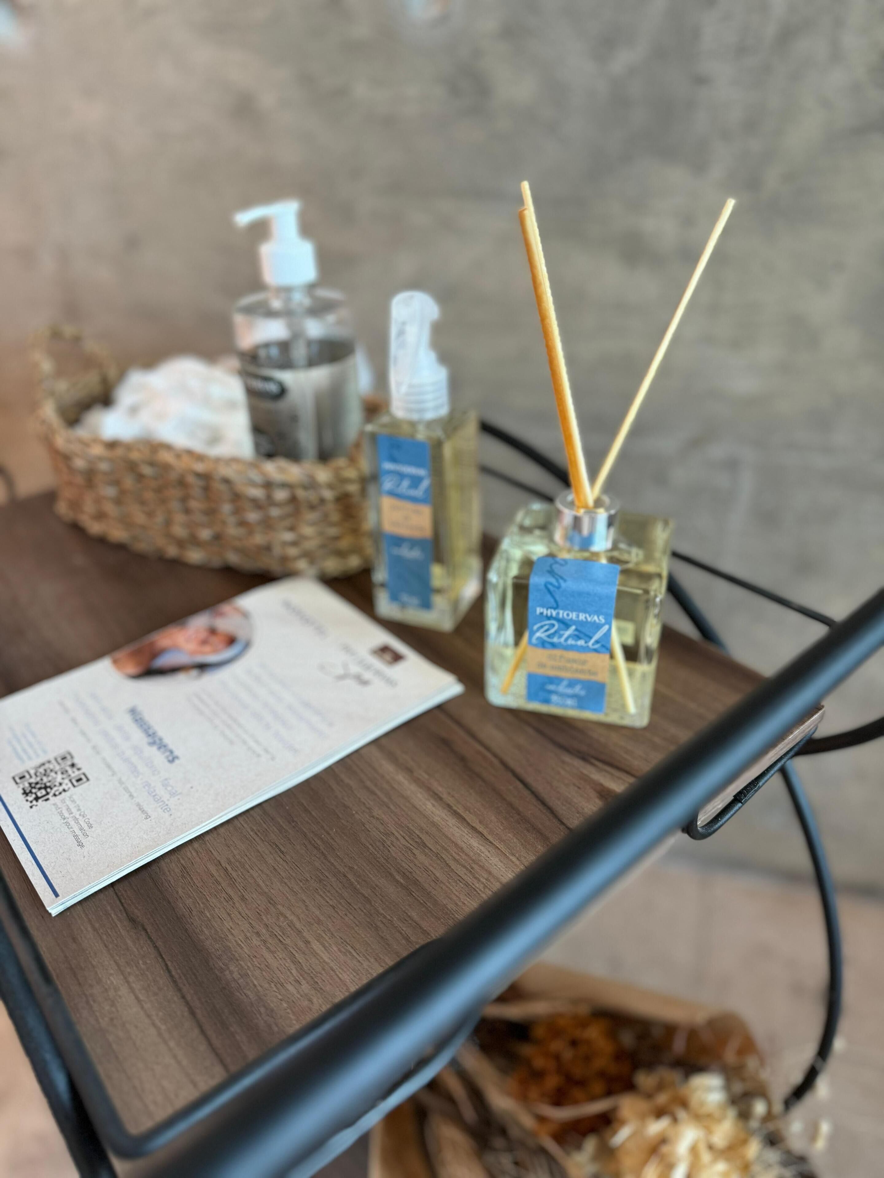 Uma mesa de madeira com um folheto, um frasco de álcool em gel, um óleo essencial e um difusor de vime.