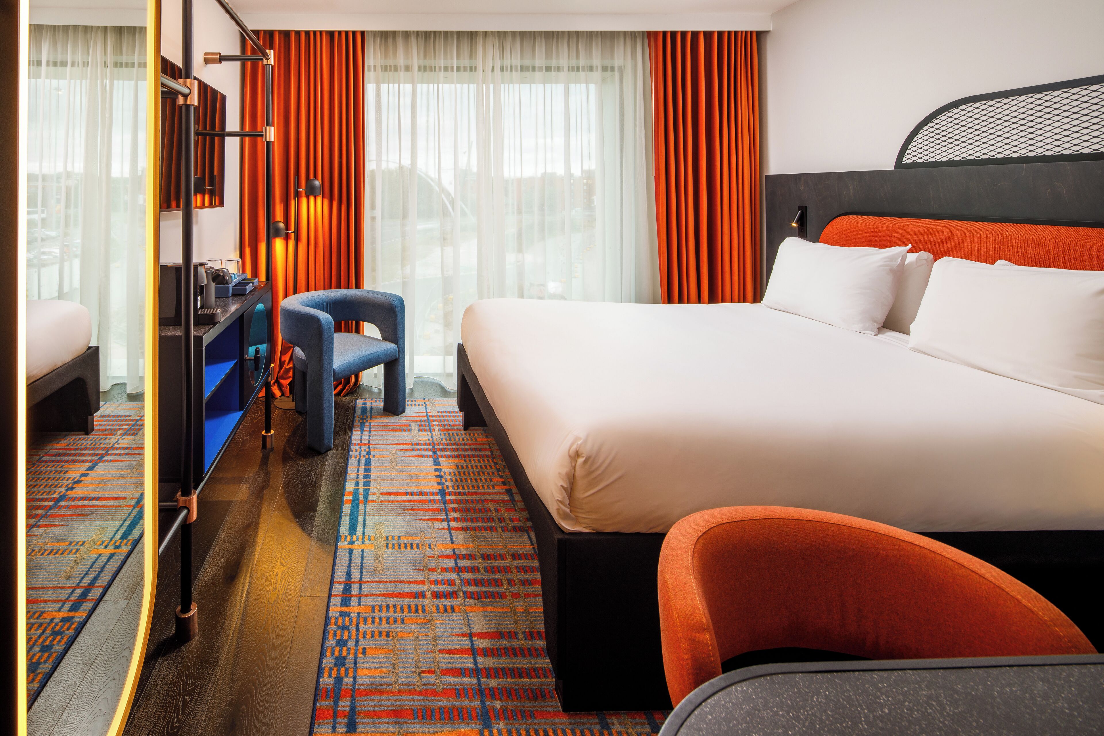 Quarto de hotel com uma cama grande, uma escrivaninha com cadeira, um sofá pequeno e um tapete estampado.