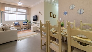 Uma sala de estar com sofá, cadeiras, mesa de jantar posta para quatro pessoas, televisão e lustre.