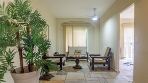 Sala ampla com uma planta em vaso grande, ventilador de teto e vista pela janela com cortinas.