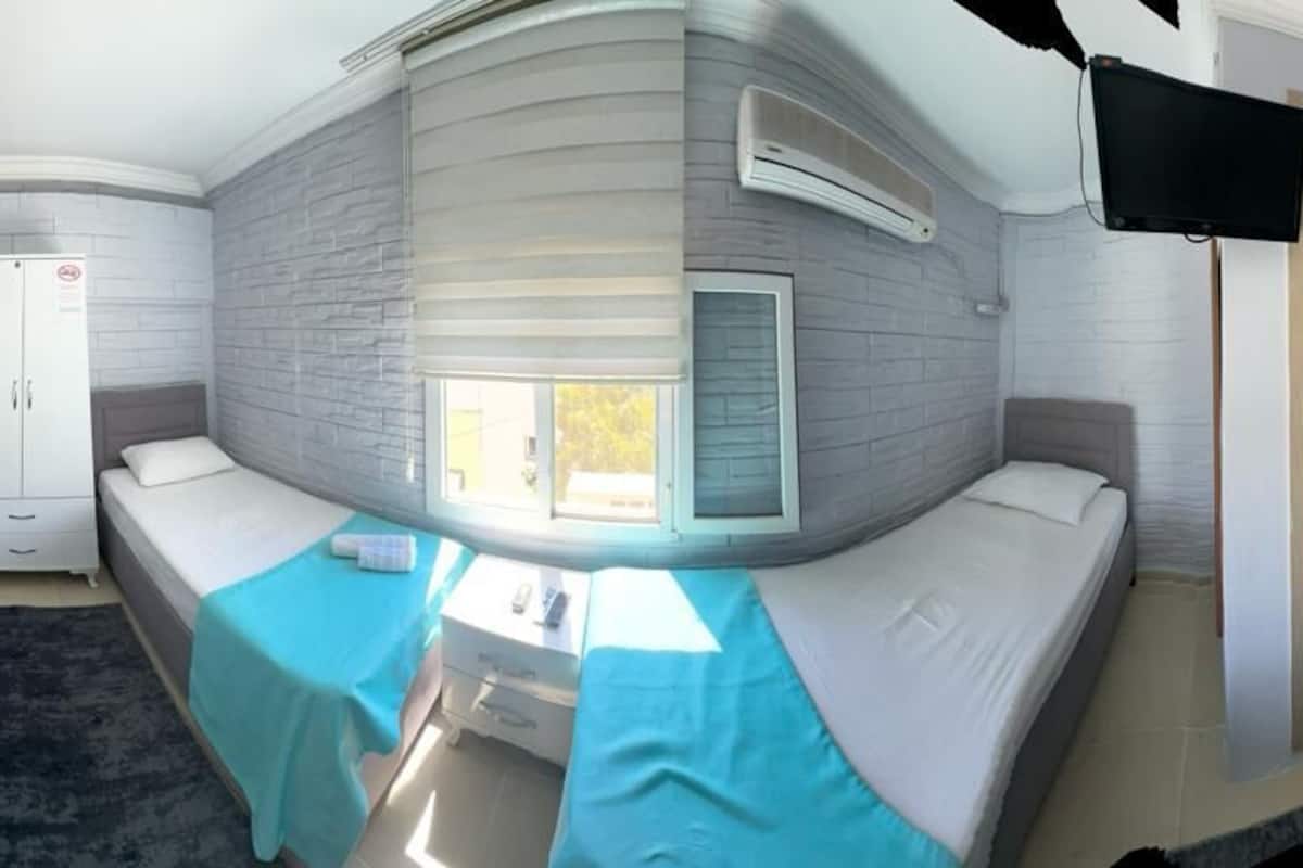 Quarto de hotel compacto com duas camas de solteiro, um pequeno espaço de estar, um guarda-roupa e uma televisão.