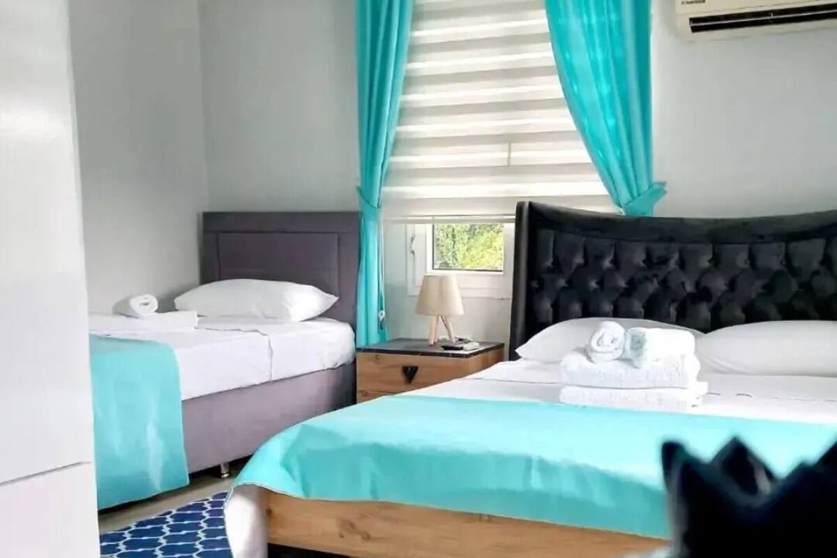 Quarto com duas camas, uma janela com cortinas, mesa de cabeceira com abajur e ar-condicionado fixado na parede.