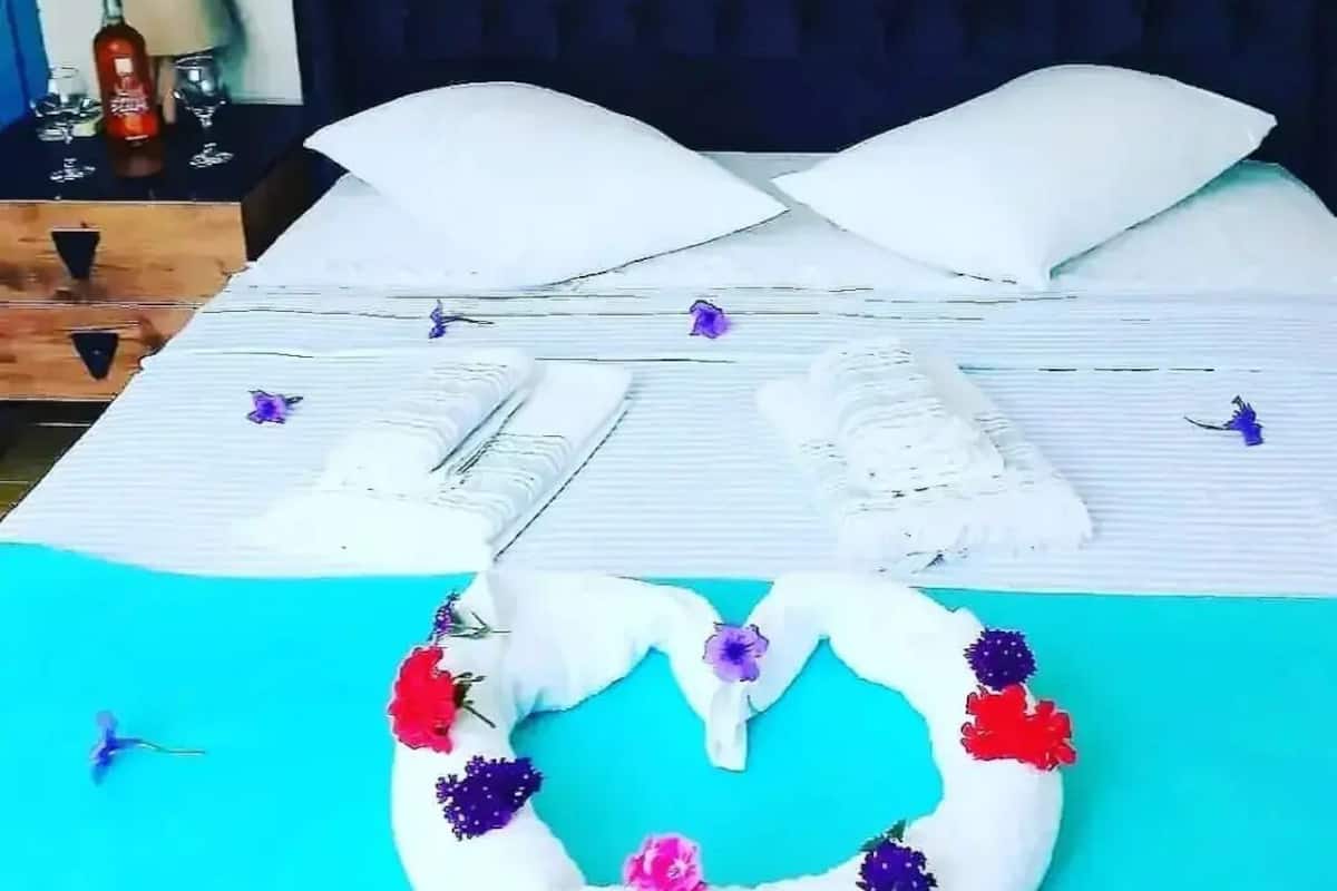 Cama bem arrumada com almofadas brancas e colcha azul, mesa de cabeceira de madeira com uma garrafa e copos, e parede com padrão azul escuro.
