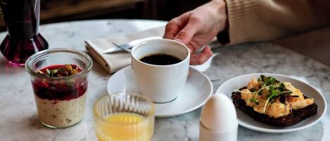 Mesa de café da manhã com xícara de café, copo de suco de laranja, tigela de mingau e prato com ovos mexidos e folhas verdes.