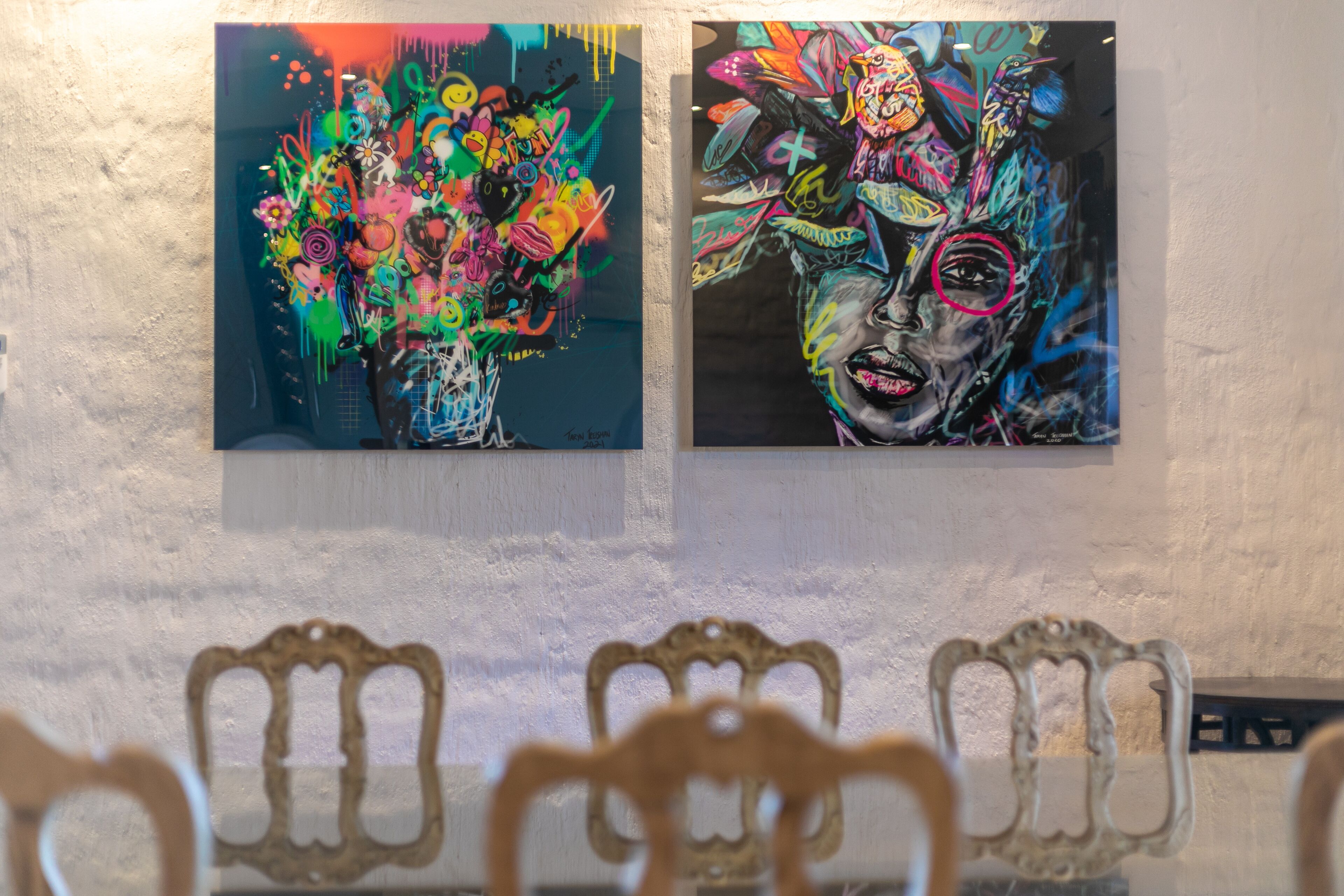 Duas pinturas vibrantes numa parede, com uma cadeira rústica em primeiro plano.