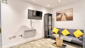 Uma sala de estar moderna com uma TV de tela plana na parede, um sofá cinza com almofadas amarelas, uma mesa de centro de vidro e uma mesinha de cabeceira de madeira.