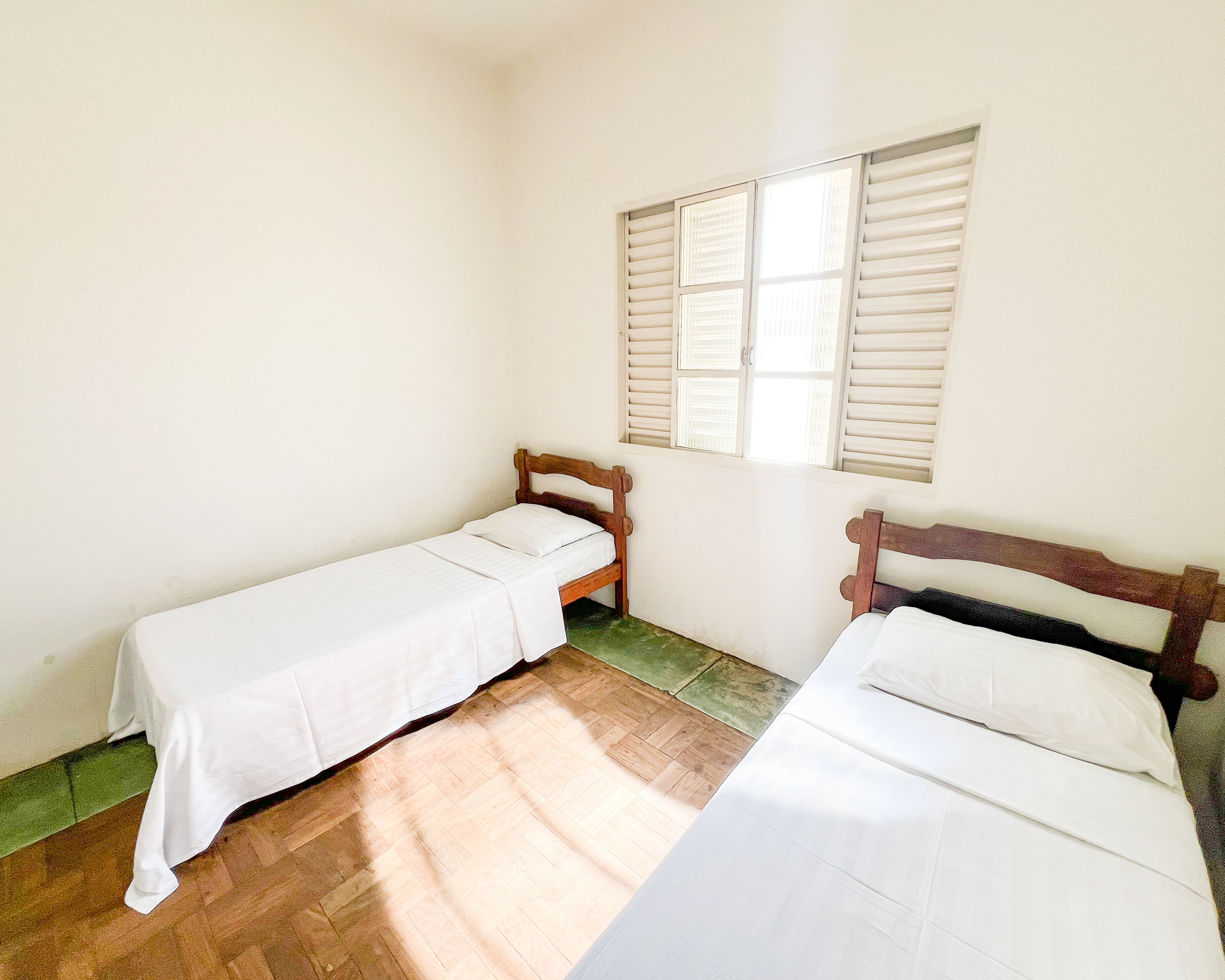 Um quarto pequeno e simples, com duas camas individuais, uma janela com persianas e uma parede sem adornos.
