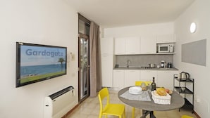 Cozinha compacta com armários brancos, micro-ondas e pia. Há uma mesa de jantar com cadeiras amarelas e uma cesta de pão. Uma televisão na parede exibe uma cena de praia.