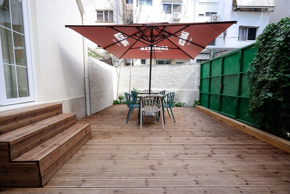 Um deck de madeira com mesa, cadeiras e um guarda-sol, além de uma cerca verde.