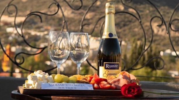 Uma mesa com uma garrafa de vinho Riunite Esprume, dois copos vazios, um prato de queijo e uma rosa numa varanda com vista para as montanhas.