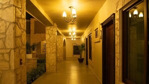 Um corredor com paredes de pedra, chão ladrilhado e um candelabro. Há plantas em vasos e uma porta no lado direito.