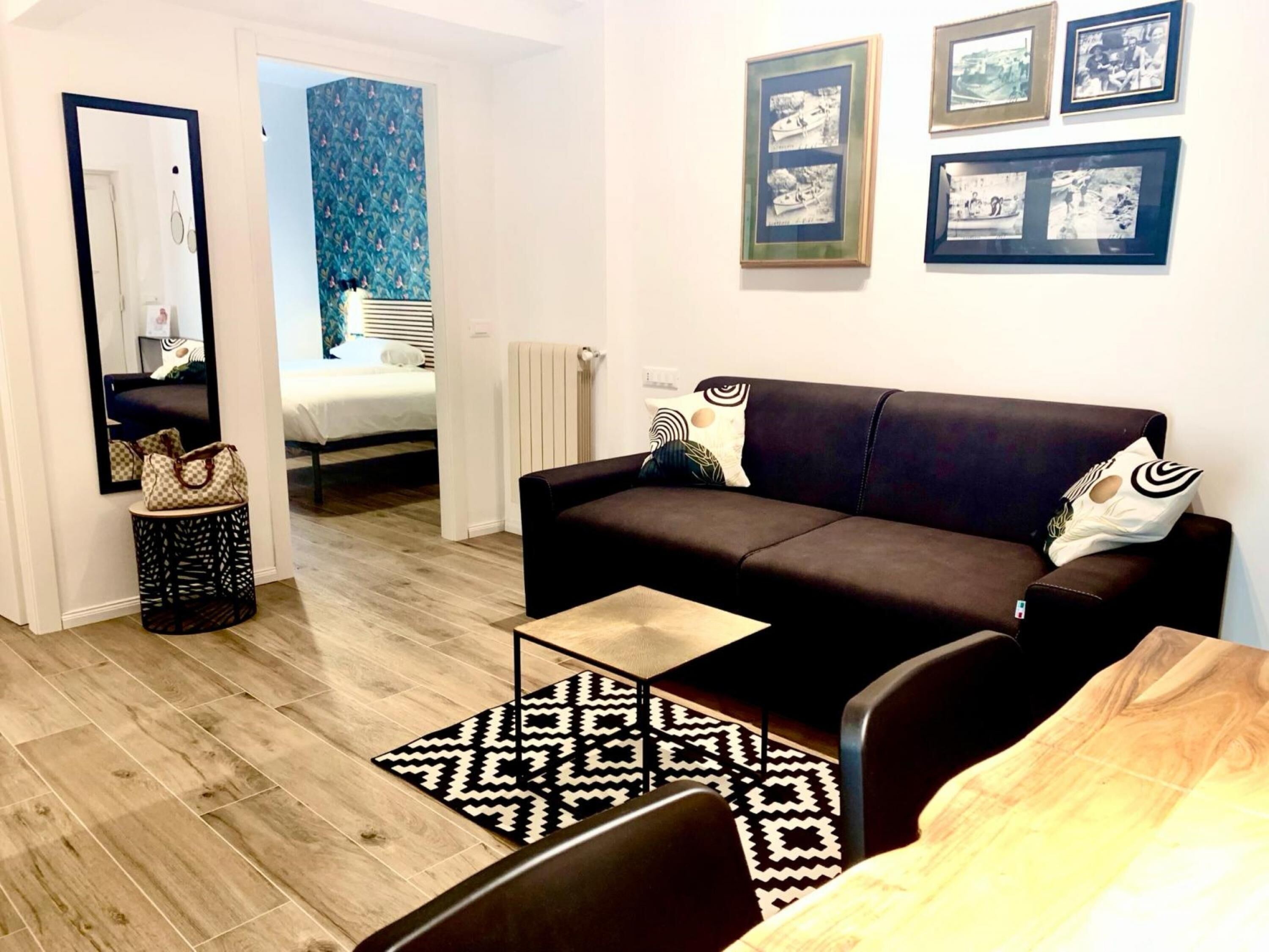 Uma sala de estar moderna com um sofá marrom escuro, um tapete estampado em preto e branco e uma mesa de jantar de madeira. Há fotografias emolduradas na parede e um espelho.