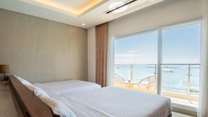 Quarto de hotel com uma cama grande, uma mesa com telefone e vista para o mar por grandes janelas.