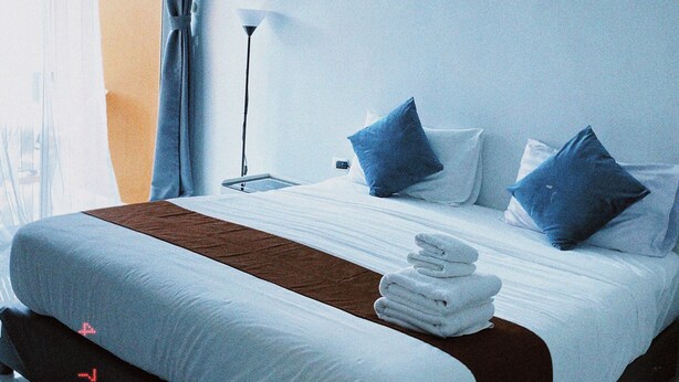 Quarto de hotel com cama, almofadas, abajur e janela com cortinas.