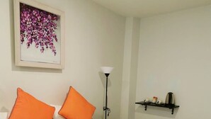 Cama bem arrumada com roupa de cama branca e duas almofadas laranjas, um cobertor listrado e colorido, uma obra de arte floral emoldurada na parede, um abajur e uma pequena prateleira com objetos.