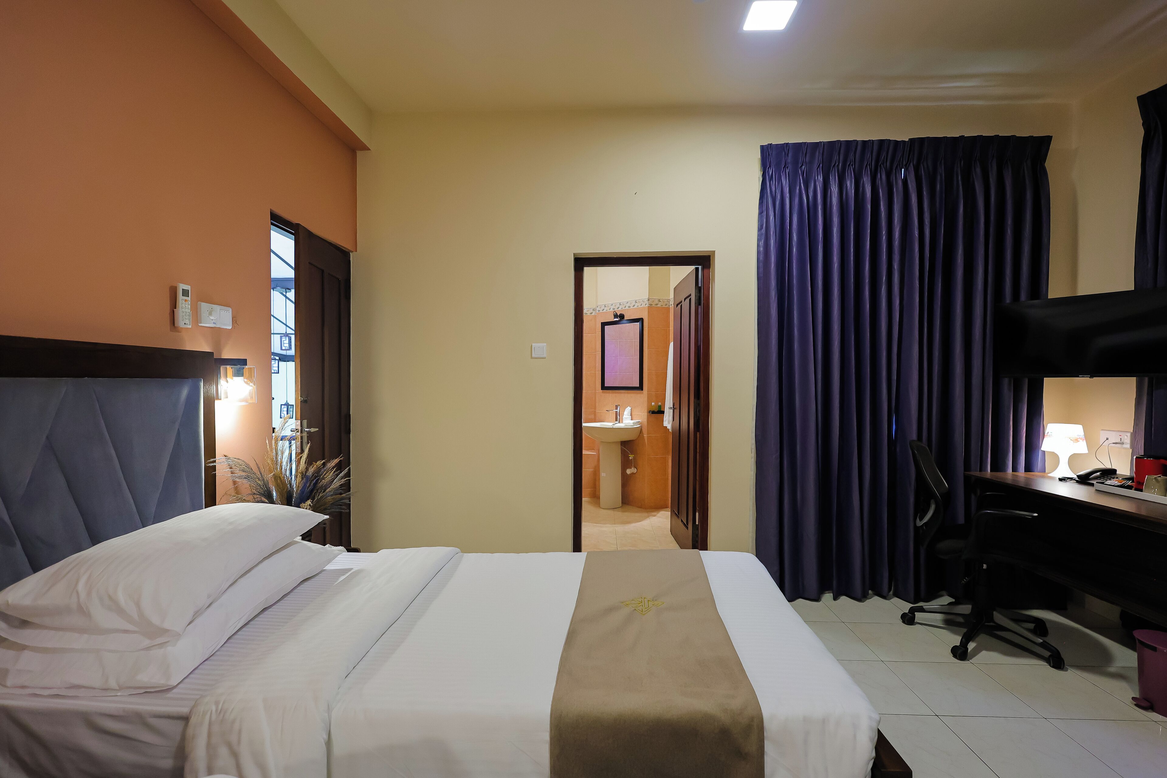 Quarto de hotel com cama, secretária e casa de banho visível através de uma porta aberta.