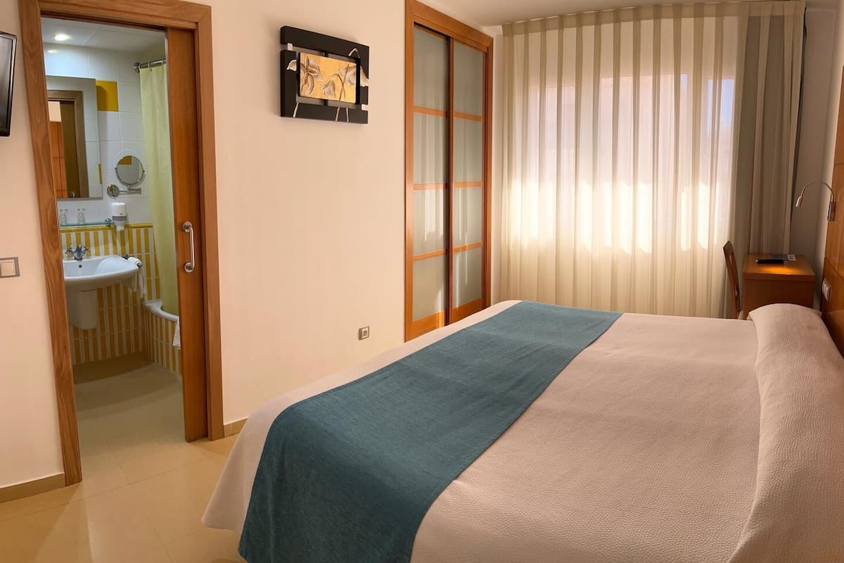 Quarto de hotel com uma cama, banheiro com pia e espelho, e um guarda-roupa com porta deslizante.