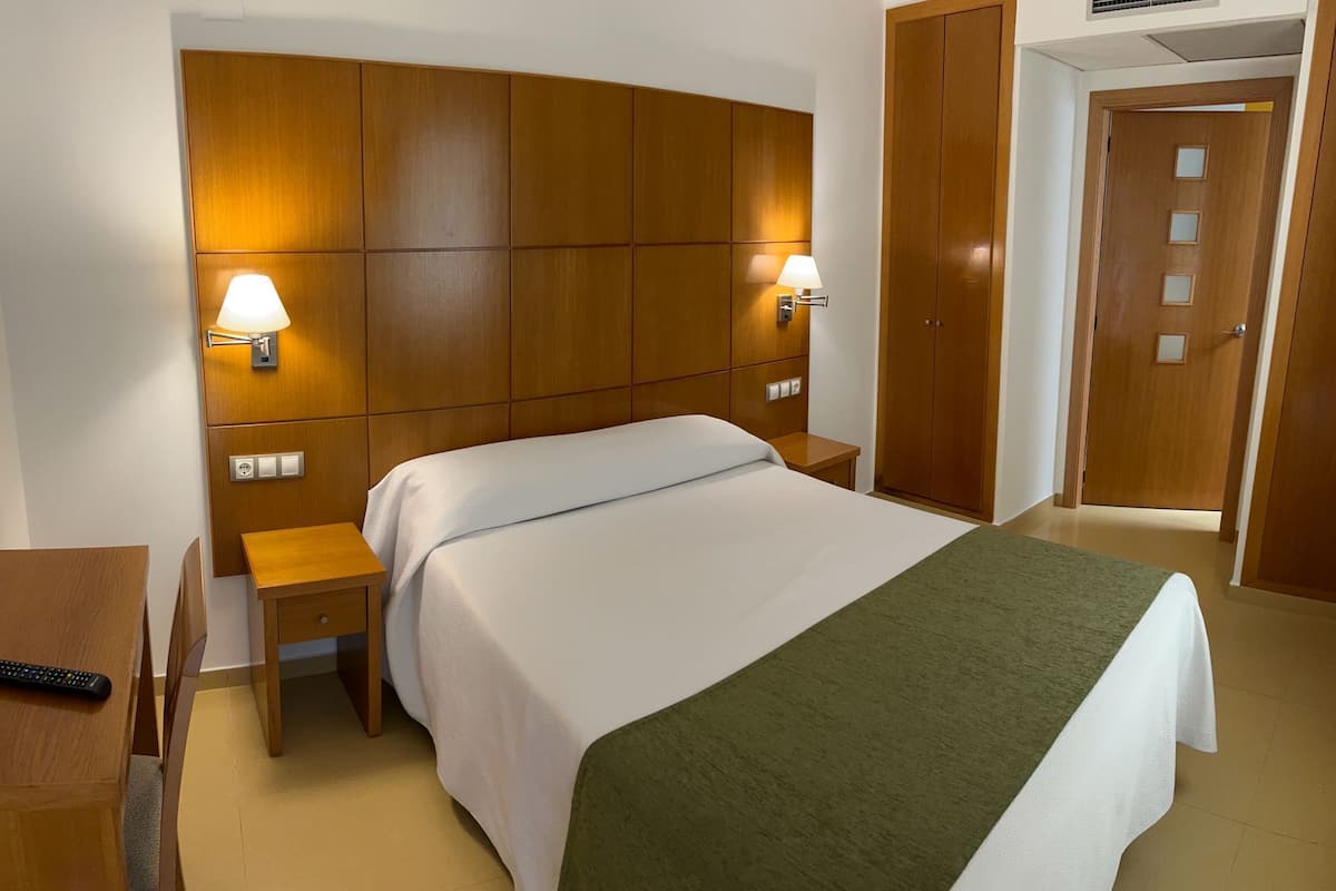 Quarto de hotel com cabeceira de madeira, duas luminárias de apoio, cama com lençol branco, manta verde, mesa de cabeceira de madeira, controle remoto e guarda-roupa de madeira.
