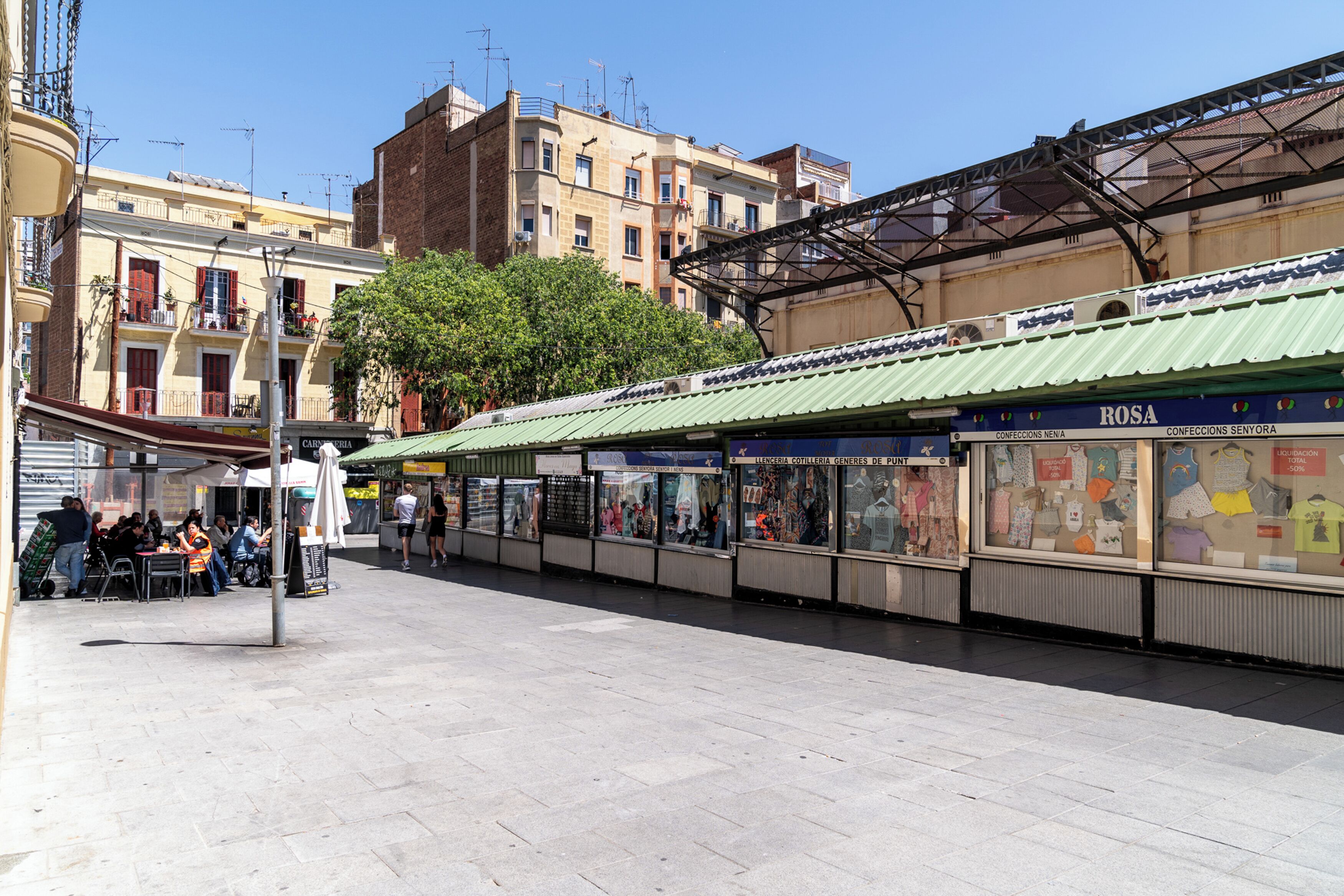 Uma praça pública com mesas ao ar livre, área coberta com lojas e edifícios ao fundo.
