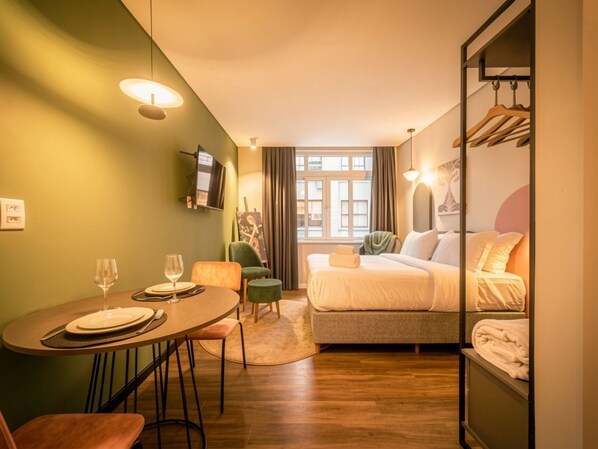 Quarto de hotel com cama, mesa de jantar e cadeiras, televisão fixada na parede e janela com cortinas.