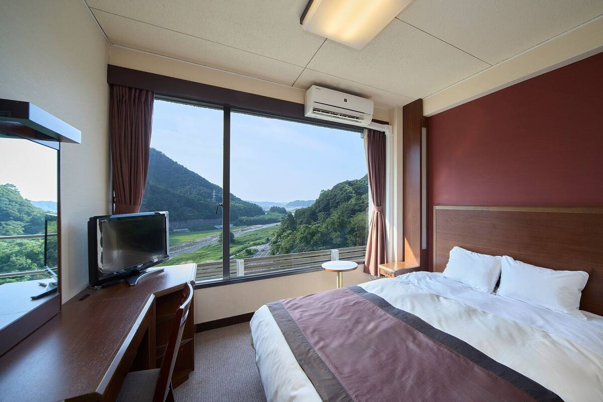 Quarto de hotel com uma cama, uma escrivaninha com televisão e uma janela com vista para um vale.