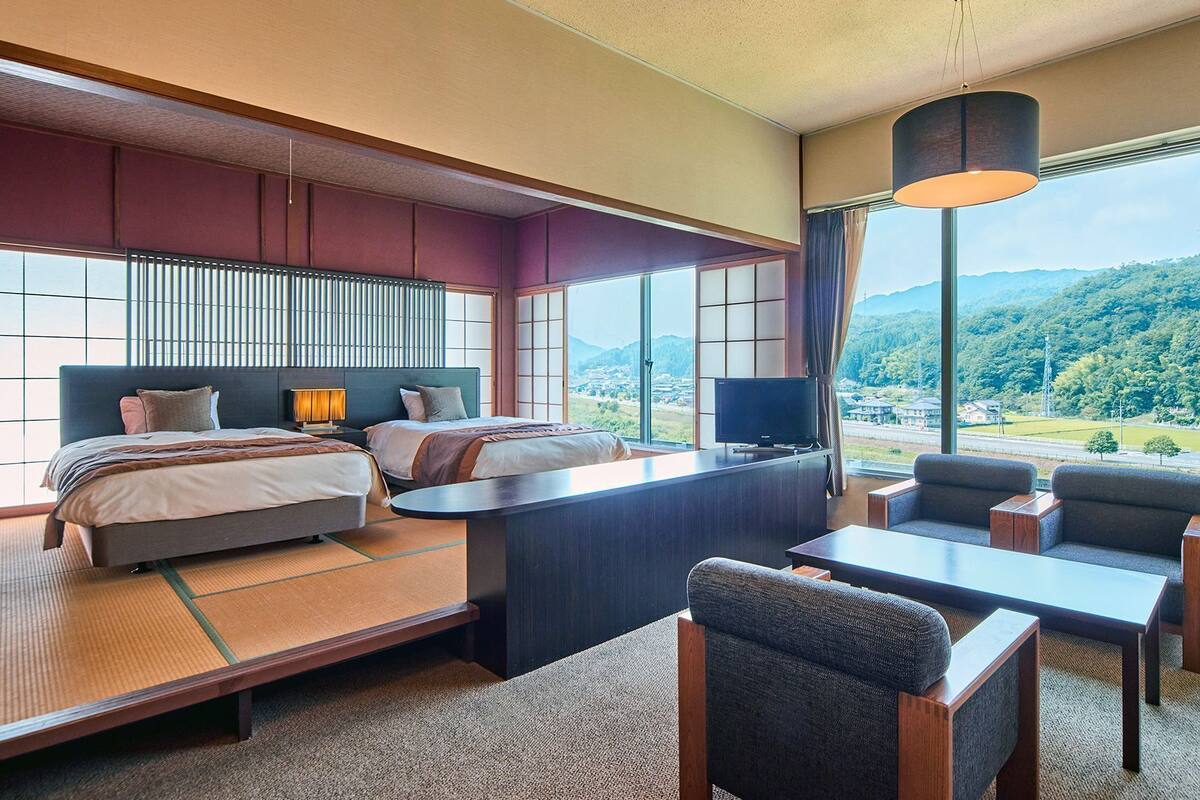 Quarto de hotel com duas camas, um sofá, uma TV e vista para uma paisagem montanhosa.