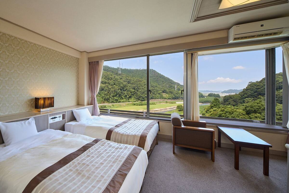 Quarto de hotel com duas camas, uma escrivaninha, uma cadeira e grandes janelas com vista para uma paisagem arborizada.