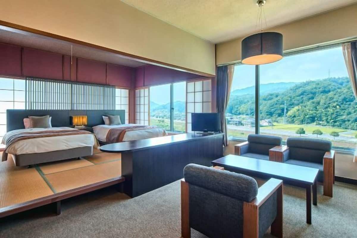 Quarto de hotel com cama, sofá e cadeiras, janelas amplas com vista para uma paisagem verde.