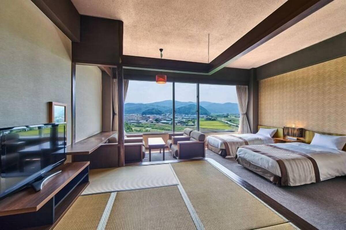 Quarto de hotel com uma janela grande, televisão de tela plana, duas camas, um sofá e uma mesa pequena.