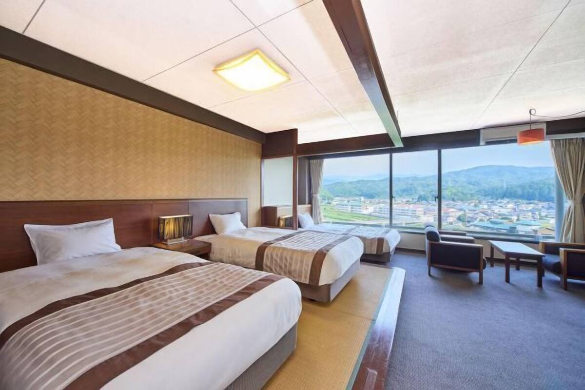 Quarto de hotel com duas camas, um sofá, uma escrivaninha e uma janela grande com vista para a cidade.