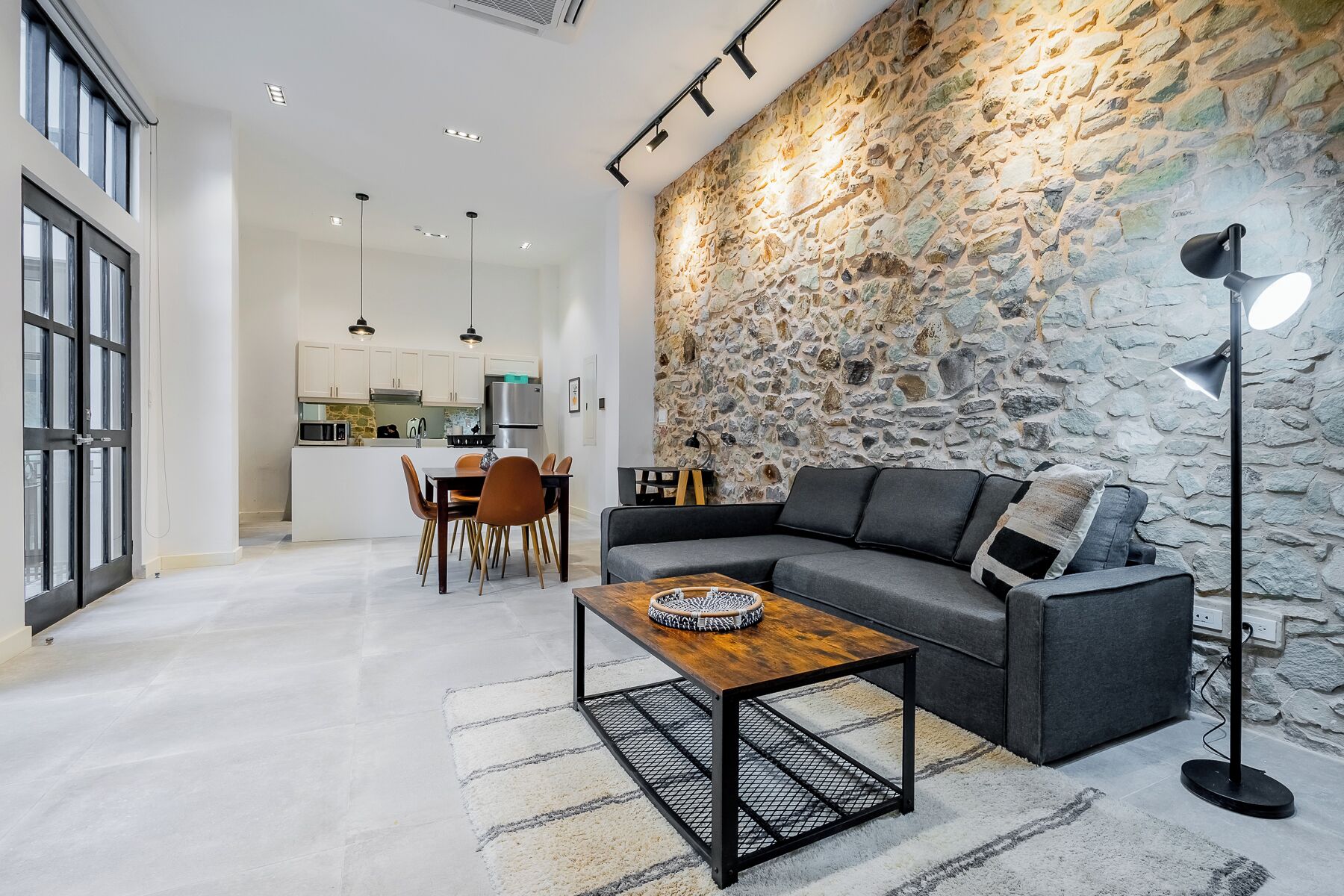Uma sala moderna com uma parede de destaque em pedra, um sofá cinza, uma mesa de centro de madeira e uma área de jantar com mesa e cadeiras brancas.