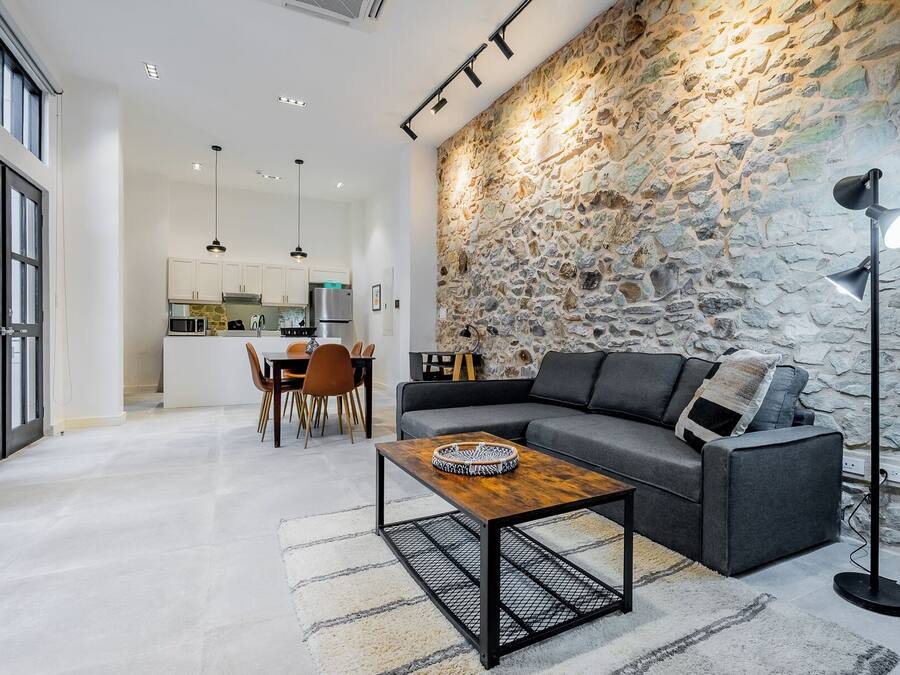 Uma sala moderna com uma parede de destaque em pedra, um sofá cinza, uma mesa de centro de madeira e uma área de jantar com mesa e cadeiras brancas.