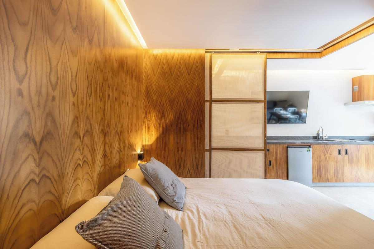 Quarto com cabeceira de madeira, cama com duas almofadas, mesa de cabeceira e televisão embutida.