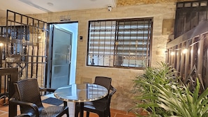 Uma área de estar externa com mesa e cadeiras de vidro, cercada por paredes de pedra e plantas em vasos.