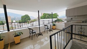 Uma varanda com vista para um horizonte urbano, incluindo edifícios e montanhas. Há uma mesa com duas cadeiras e um vaso com planta.