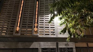 Um edifício moderno com o número '49' em destaque na fachada, grandes janelas com persianas verticais e uma árvore em primeiro plano.