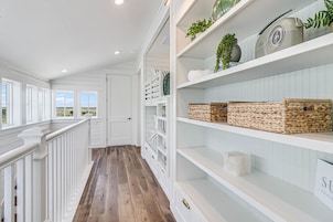 Corredor com paredes brancas, piso de madeira e um corrimão branco. Prateleiras exibem cestos de vime e objetos decorativos.