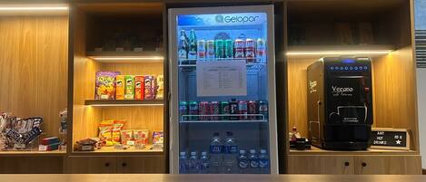 Uma máquina de vendas automática com lanches e bebidas, identificada como 'Gelopar', e uma máquina de café com a marca 'Verano cafe premium', em um ambiente de hotel.