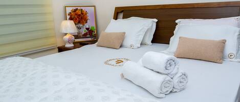 Cama bem arrumada com lençóis brancos e um edredom estampado, cabeceira de madeira e mesa de cabeceira com uma luminária e um quadro emoldurado.