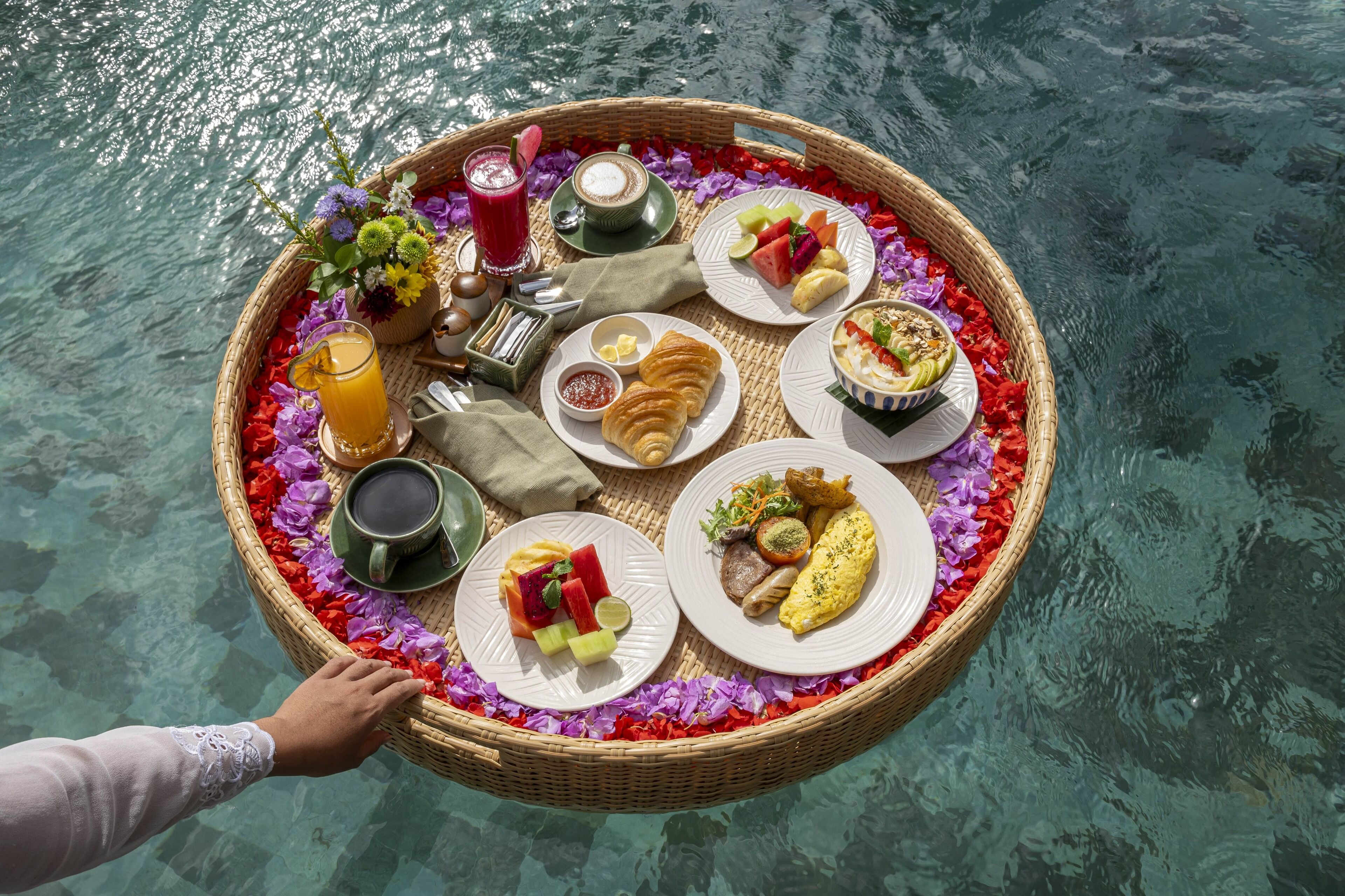 Bandeja redonda de vime com diversos itens de café da manhã, incluindo frutas, doces e bebidas, colocada sobre a água.