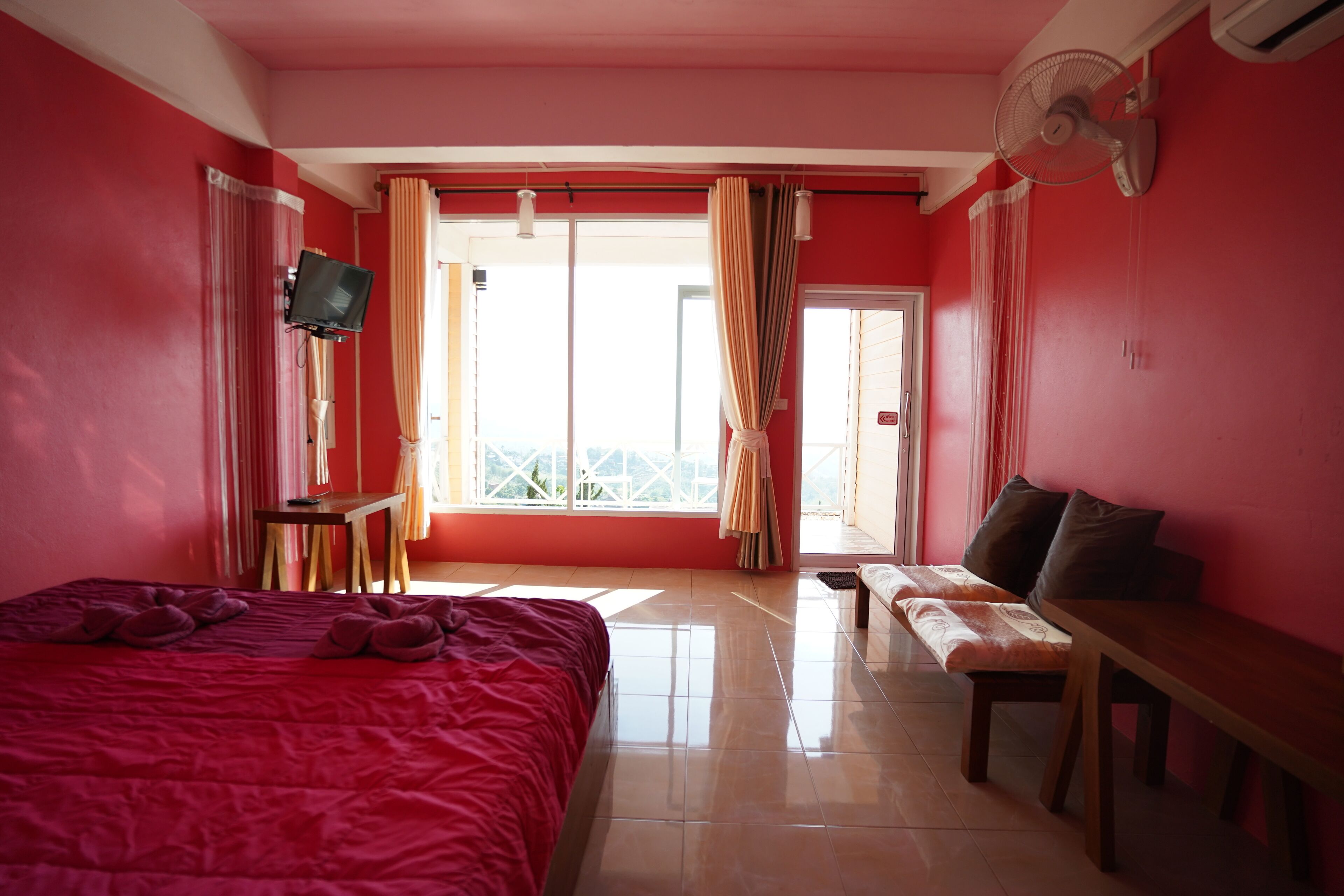 Quarto com cama, um banco, mesa, televisão e varanda com cortinas.